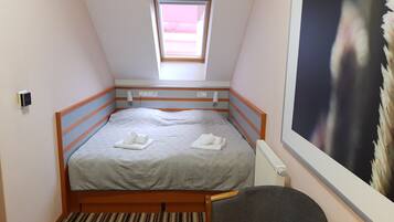 Quarto pequeno com cama de solteiro, teto inclinado, janela, radiador e uma cadeira.