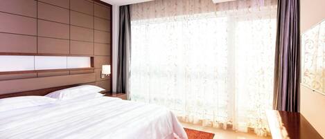 Quarto de hotel com uma cama grande, cabeceira de madeira, mesa de cabeceira e uma janela ampla com cortinas.