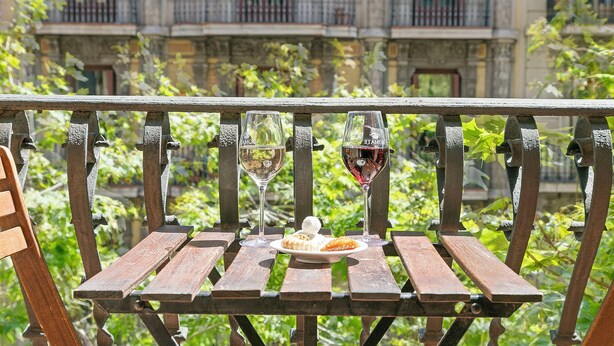 Um banco de madeira com um prato de comida e duas taças de vinho, apoiado em um corrimão de varanda com um edifício ao fundo.