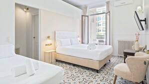 Quarto de hotel com uma cama grande, uma cama menor, uma cadeira, uma mesa de cabeceira e vista para a cidade pela janela.