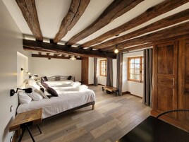Quarto com teto de madeira, cama com roupa de cama branca, mesa de cabeceira, uma cadeira e janela com cortinas.