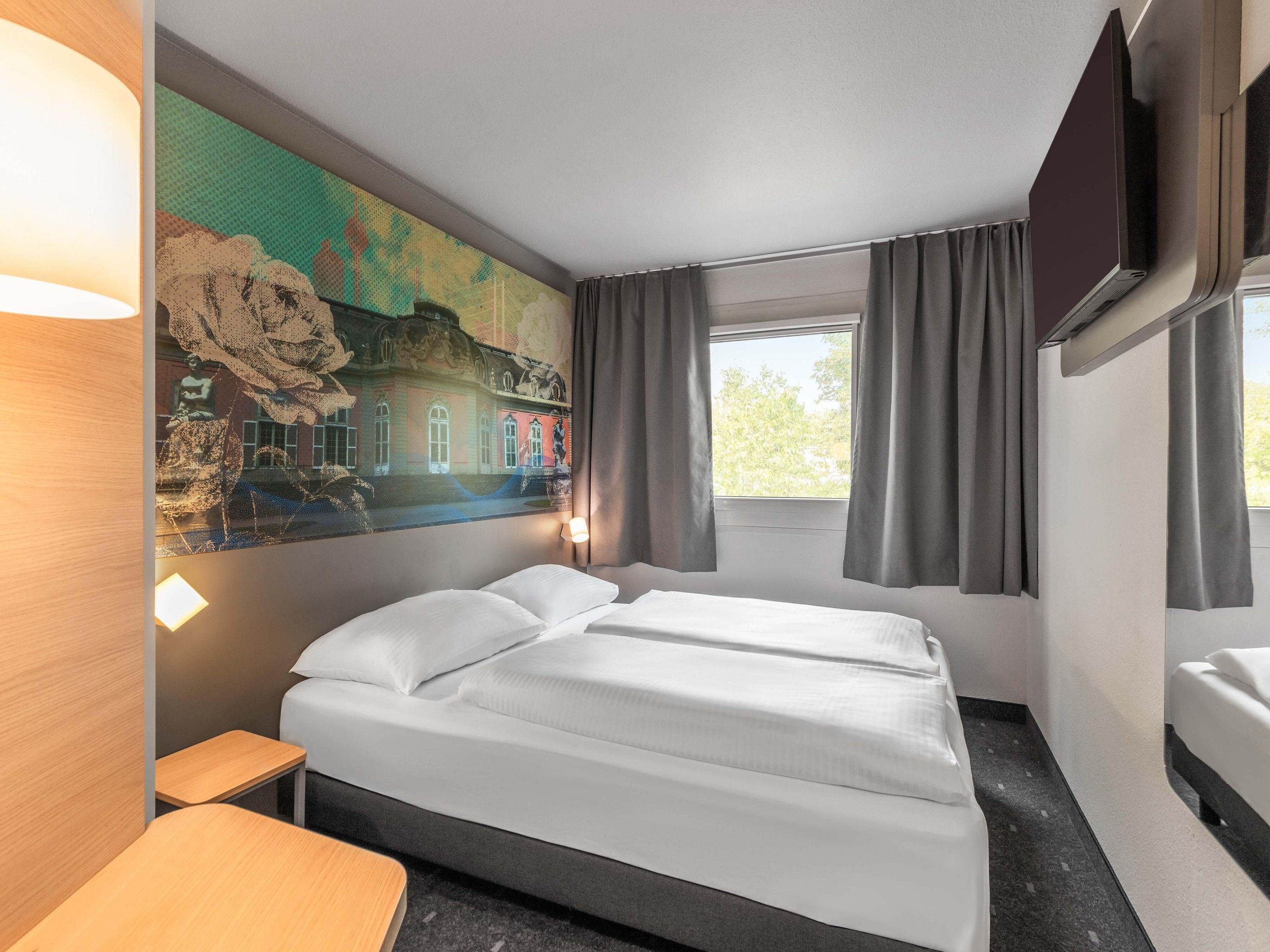 Quarto de hotel com cama, escrivaninha, janela com cortinas, televisão na parede e um quadro.