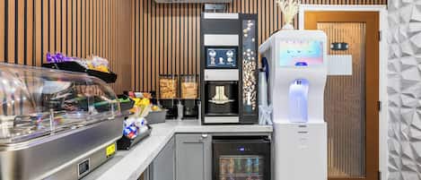 Um espaço de bebidas de serviço próprio, com máquina de gelo, cafeteira e balcão de snacks com produtos embalados.