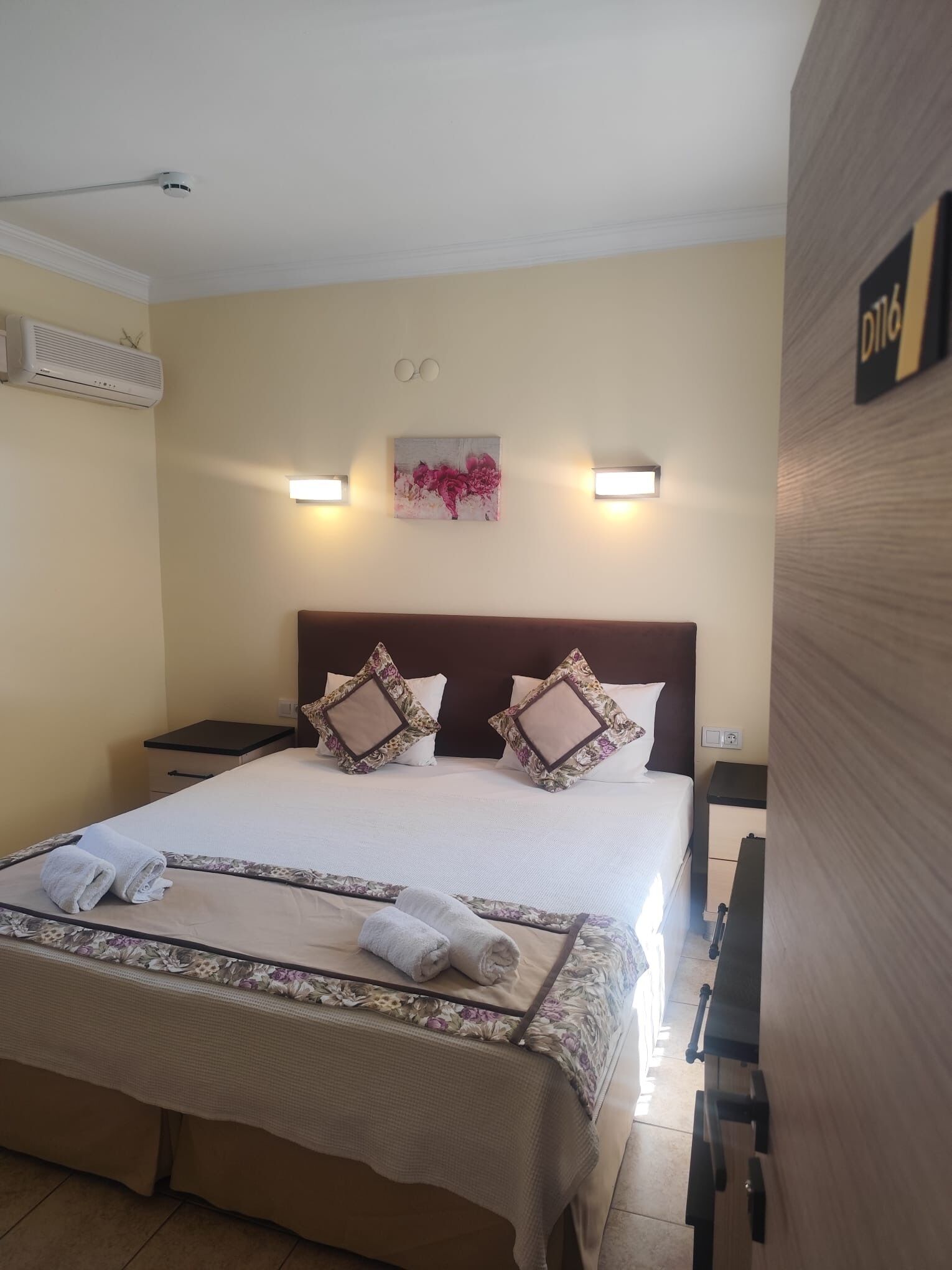 Quarto de hotel com cama, mesas de cabeceira, ar condicionado fixado na parede, um quadro e uma placa com o número '016'.