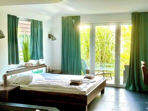 Quarto com cama, mesa de cabeceira, abajur e vista para o verde pela janela.