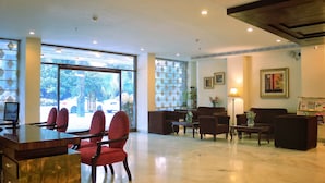 Um saguão de hotel com balcão de recepção, cadeiras vermelhas, um sofá e uma mesa de centro. Há grandes janelas com vista para o exterior.