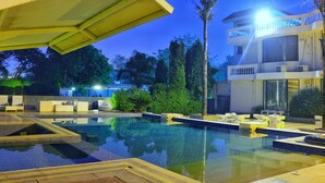Piscina com espreguiçadeiras e área coberta, cercada por vegetação e edifícios modernos.