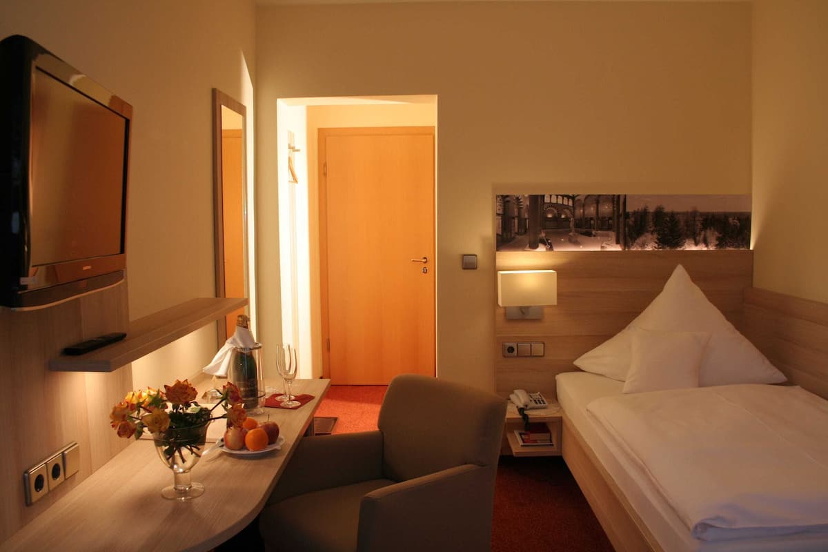 Quarto de hotel com cama, escrivaninha com luminária, televisão e pintura na parede.