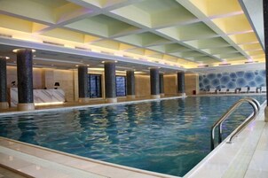 Piscina interior com piso ladrilhado, corrimão de metal e teto com padrão geométrico.