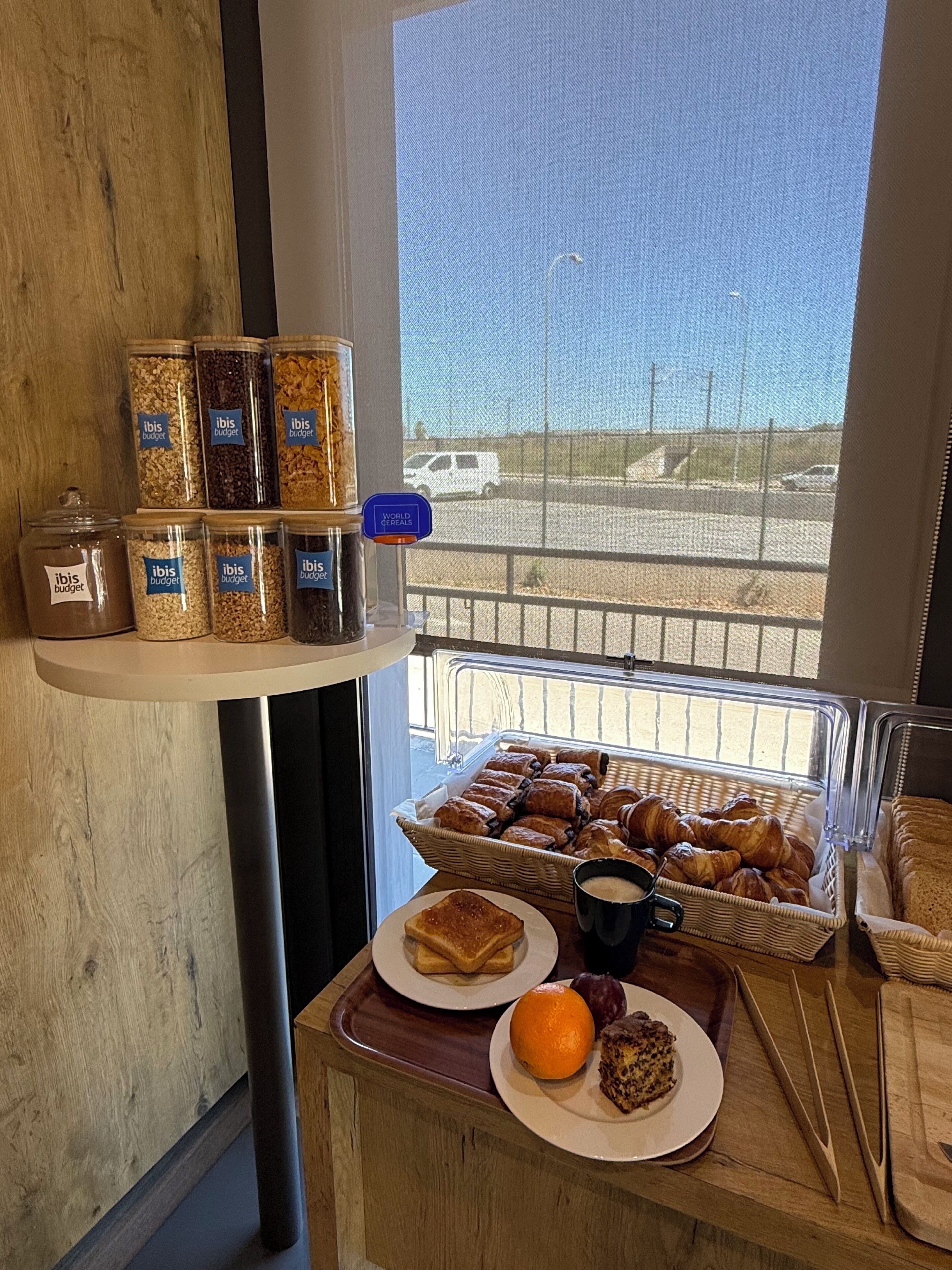 Buffet de café da manhã com cereais, pães e vista para um estacionamento através de uma janela.