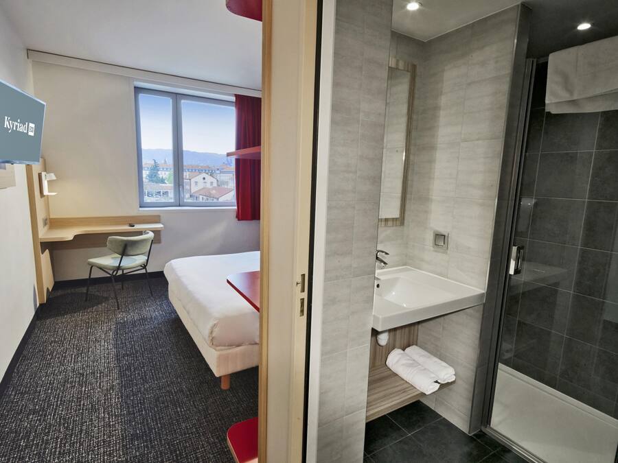 Quarto de hotel com cama, escrivaninha, cadeira e área de chuveiro.
