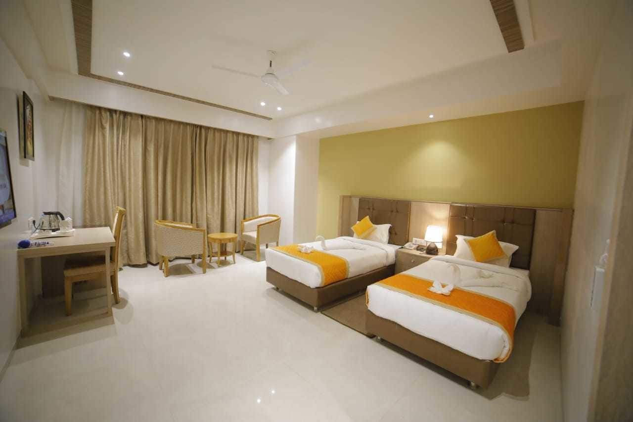 Quarto de hotel com duas camas, uma mesa, uma cadeira e uma janela grande com cortinas.