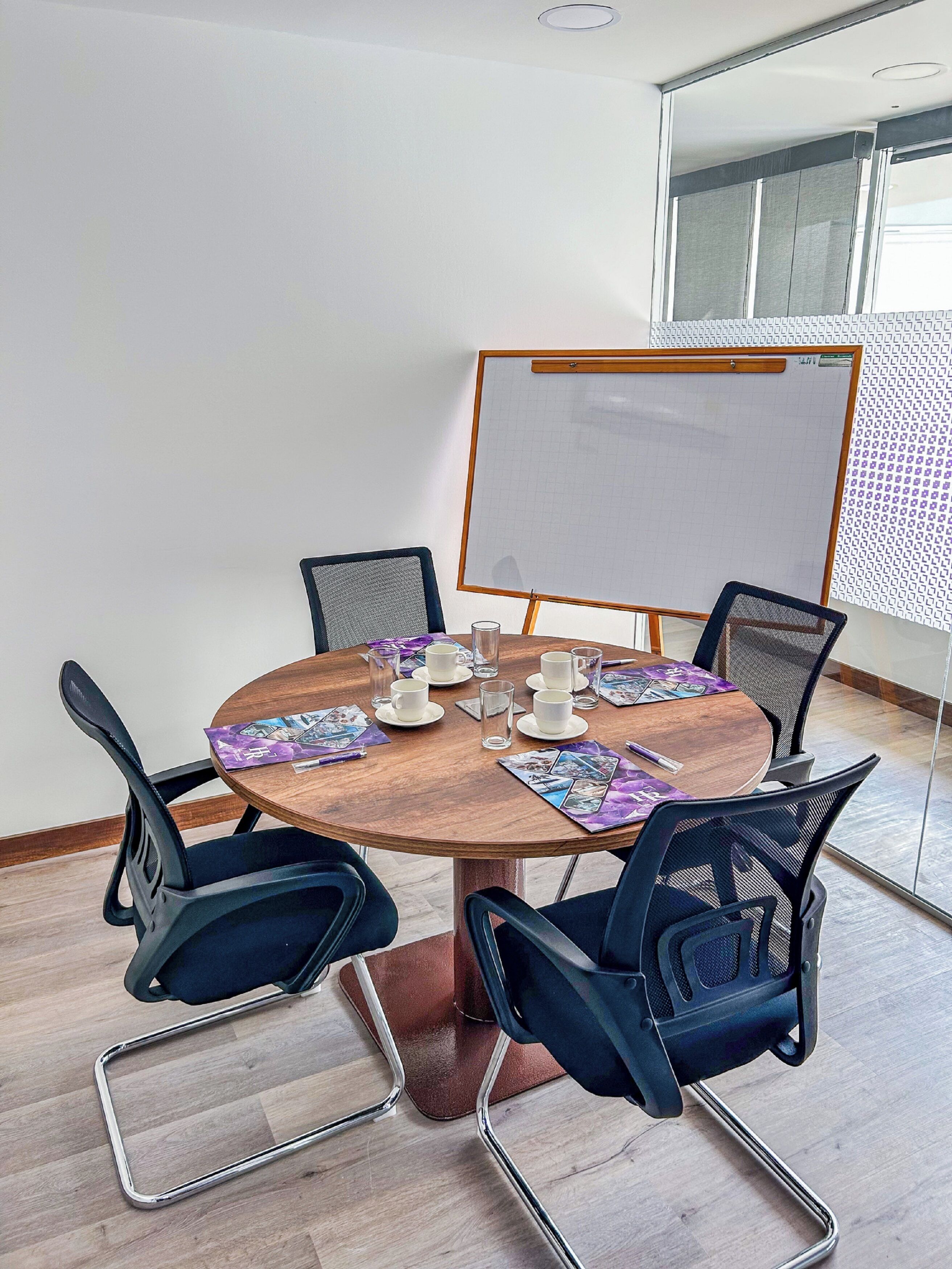 Sala de conferência com uma mesa redonda de madeira, seis cadeiras pretas, um quadro branco e um divisória de vidro.