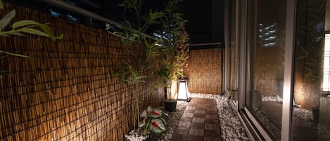 Caminho ao ar livre à noite, com parede de bambu, plantas em vasos e um candeeiro.