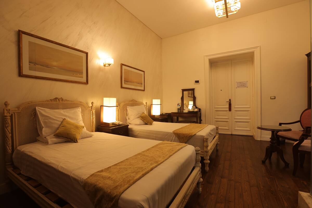 Quarto de hotel com duas camas, piso de madeira, uma mesa pequena e uma cadeira.