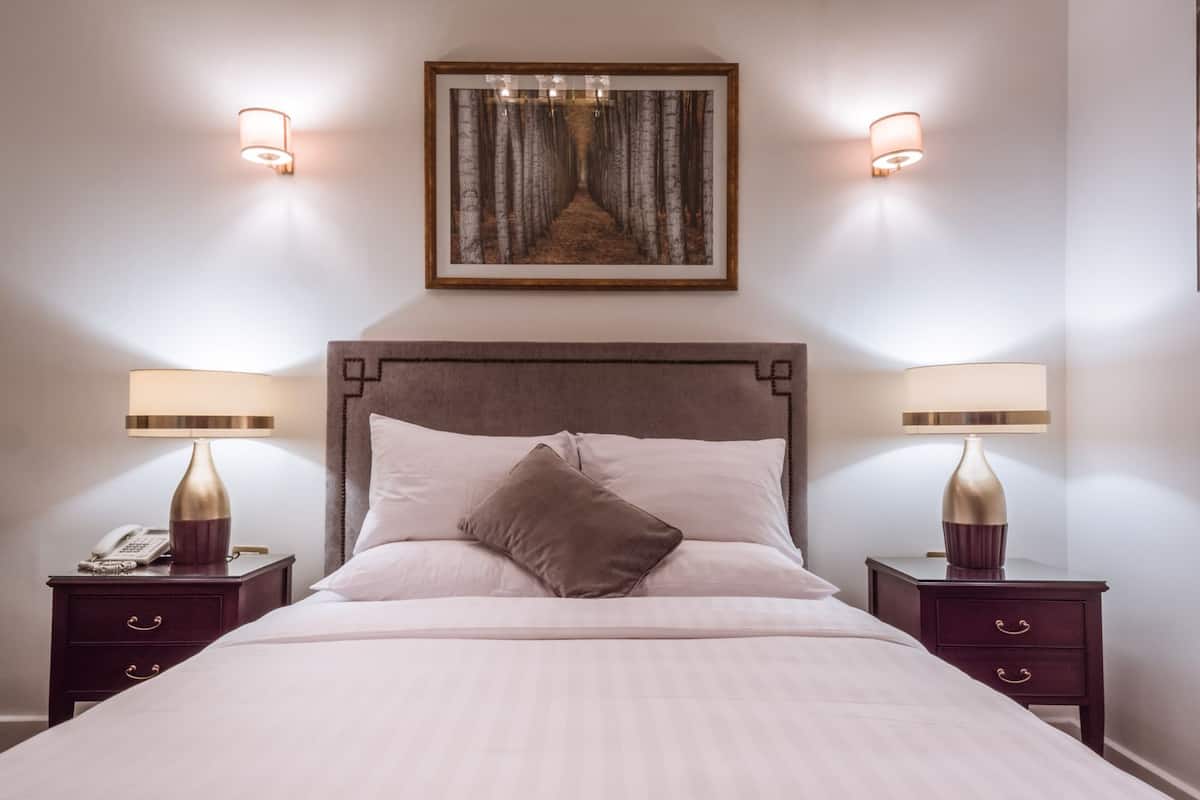 Uma cama bem arrumada com cabeceira, duas mesas de cabeceira com abajures e um quadro pendurado acima da cama.