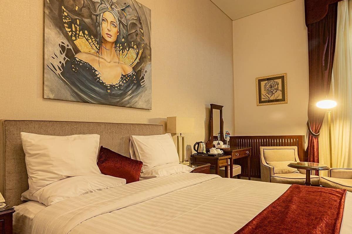 Quarto de hotel com cama grande, escrivaninha, cadeira, luminária e um quadro na parede.