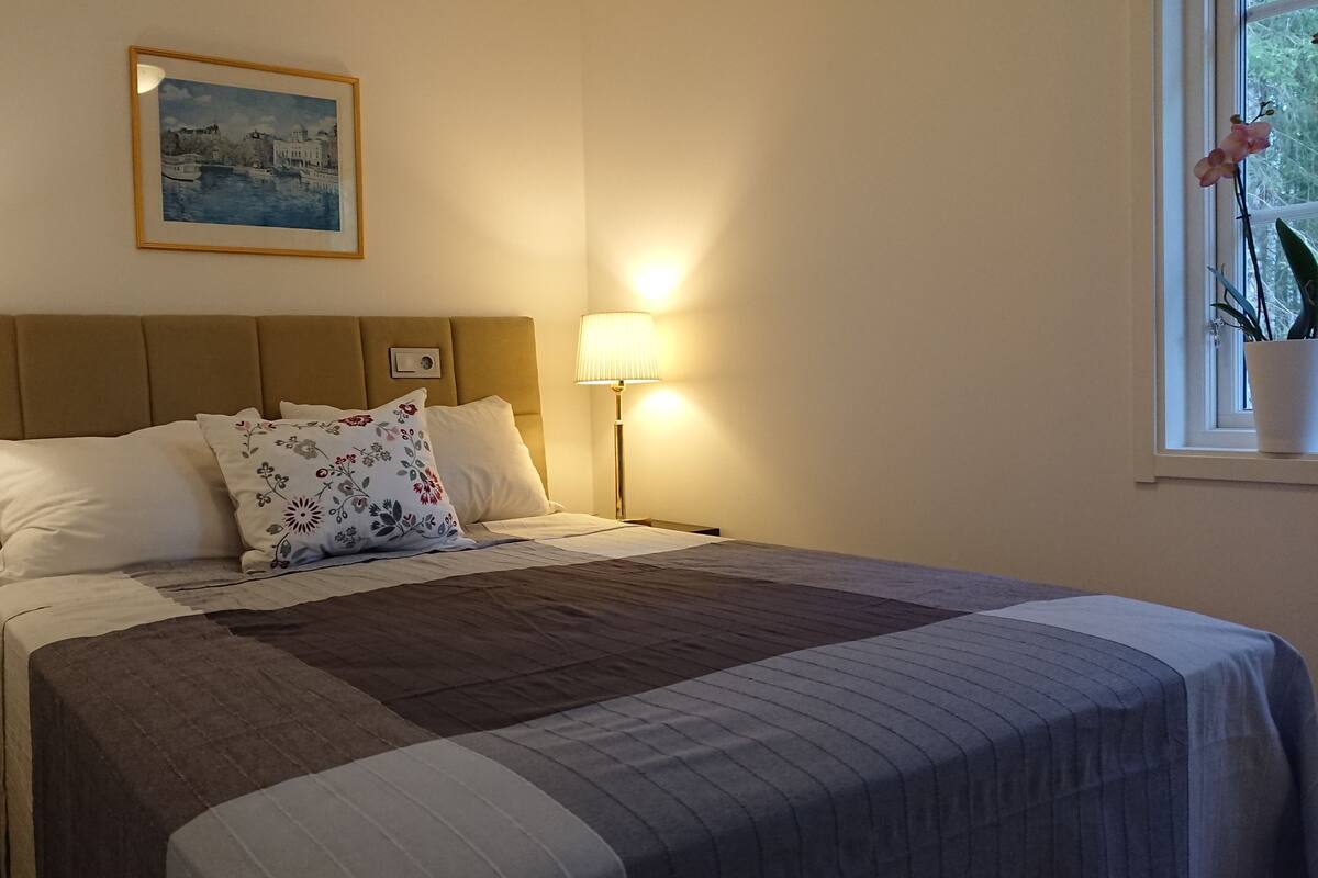 Cama bem arrumada com cabeceira, duas luminárias de apoio, almofada floral e quadro na parede.