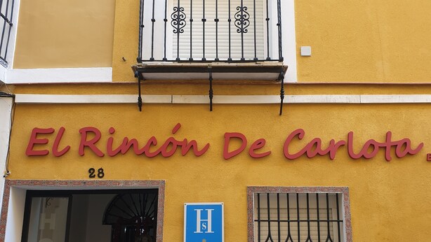 Exterior do El Rincon De Carla, um hotel com fachada amarela, uma varanda com grades de ferro preto e um letreiro azul indicando que é um hotel.