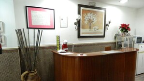 Bancada de recepção com acabamento em madeira, um vaso com gravetos, um certificado emoldurado e uma pintura floral na parede.