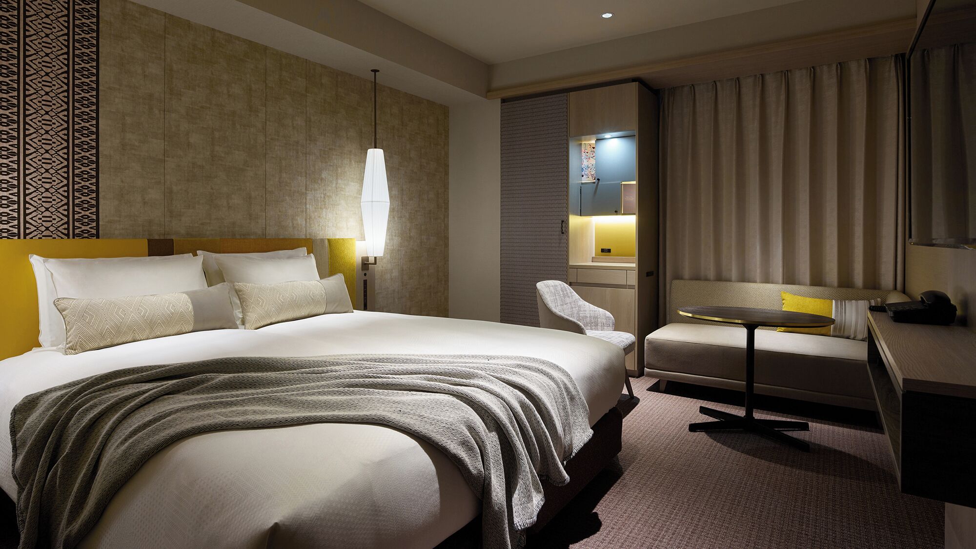 Quarto de hotel com uma cama grande, uma escrivaninha com cadeira, uma luminária e vista para a cidade.