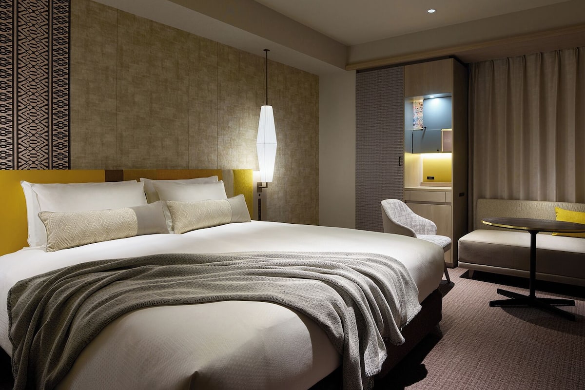 Quarto de hotel com uma cama grande, uma escrivaninha com cadeira, uma luminária e vista para a cidade.