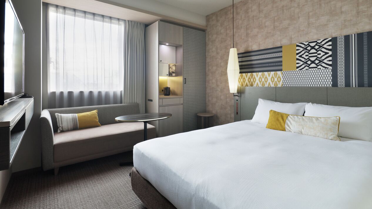 Quarto de hotel com cama, sofá, mesinha, televisão e janela com cortinas.