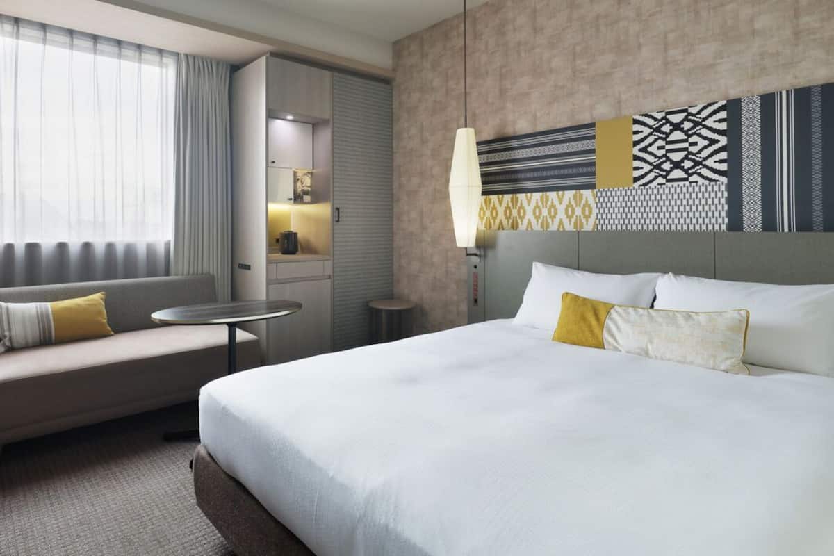 Quarto de hotel com cama, sofá, mesinha, televisão e janela com cortinas.
