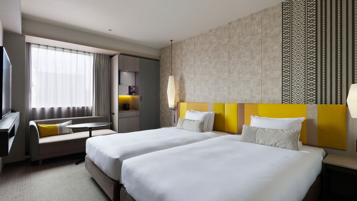 Quarto de hotel com duas camas, uma escrivaninha, um sofá e uma televisão.