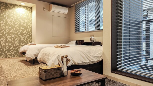 Quarto de hotel com duas camas, uma escrivaninha, uma cadeira, janela com persianas, ar condicionado e parede com padrão.