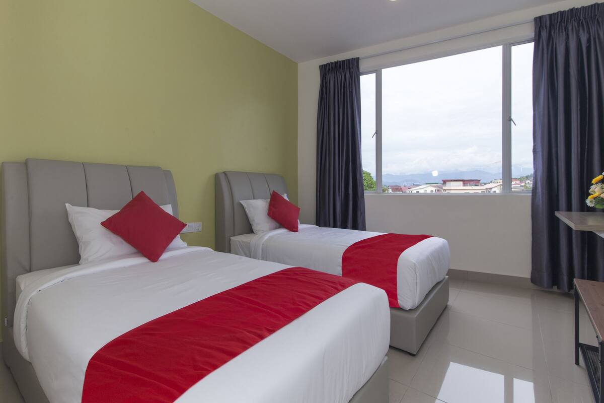 Quarto de hotel com duas camas, cada uma com roupa de cama branca e detalhes vermelhos, uma janela grande com cortinas e vista para edifícios e montanhas.