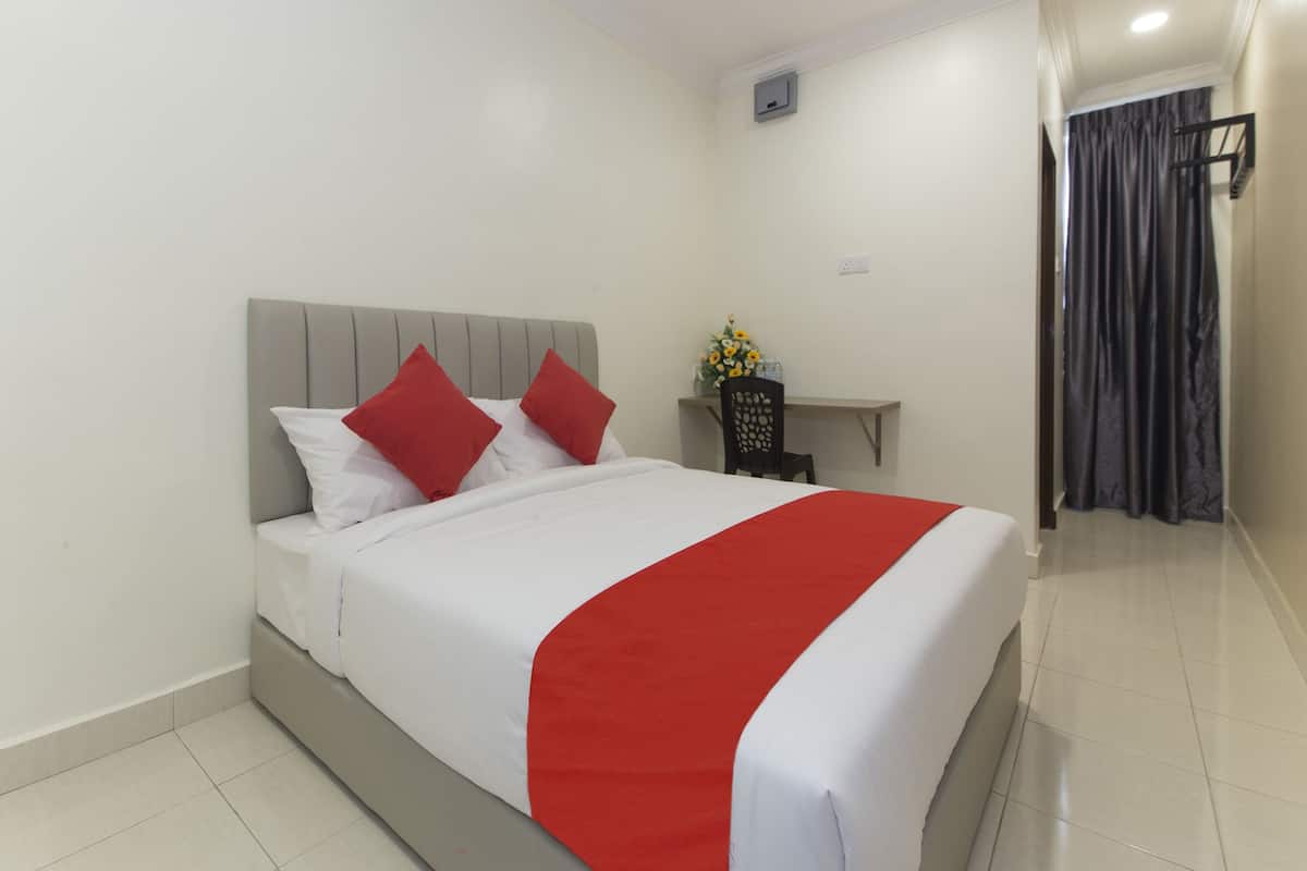 Uma cama bem arrumada com roupa de cama branca e vermelha, uma cabeceira cinzenta e uma pequena mesa com uma cadeira e um vaso de flores.