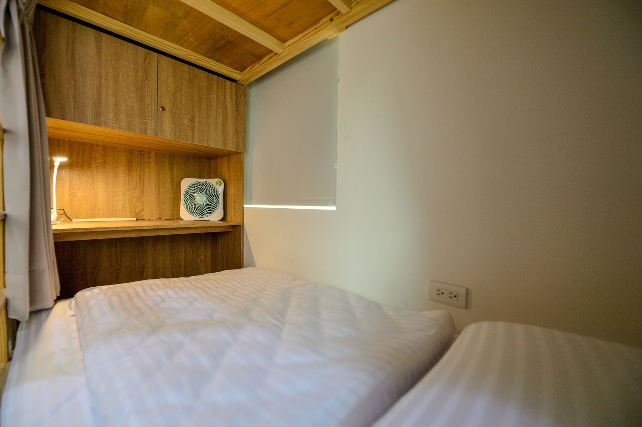 Um quarto pequeno e acolhedor, com uma cama, uma estante de madeira com um ventilador e uma janela.