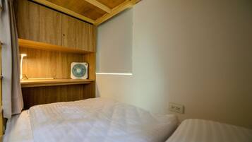 Um quarto pequeno e acolhedor, com uma cama, uma estante de madeira com um ventilador e uma janela.