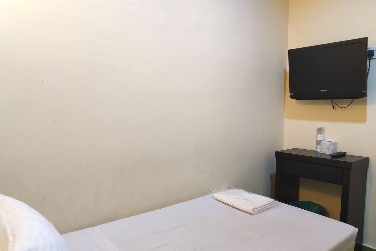 Quarto de hotel com uma cama, uma mesinha com uma garrafa e um controle remoto, e uma televisão fixada na parede.