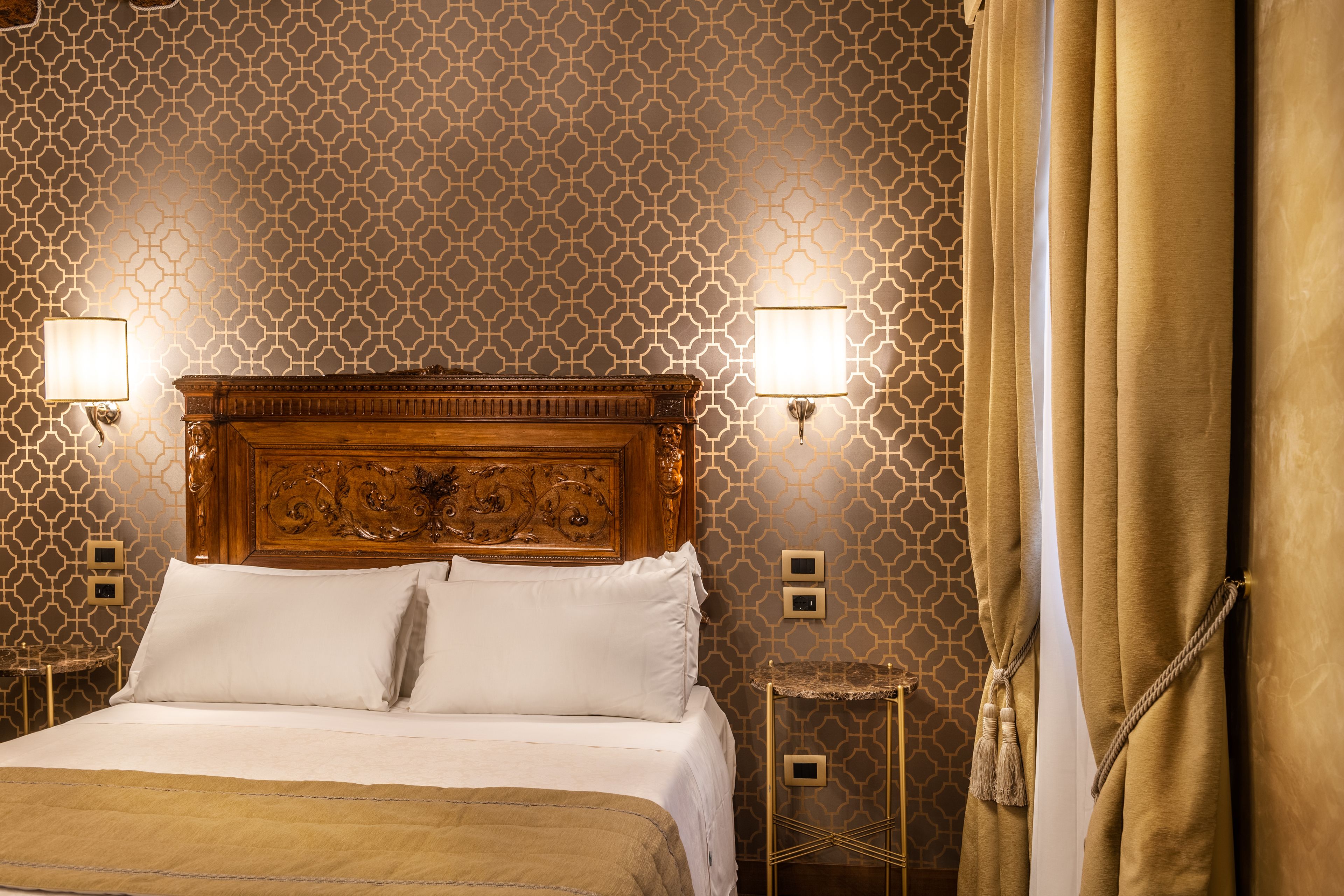 Quarto de hotel com papel de parede estampado, cabeceira de madeira, duas luminárias de cabeceira e uma cortina.