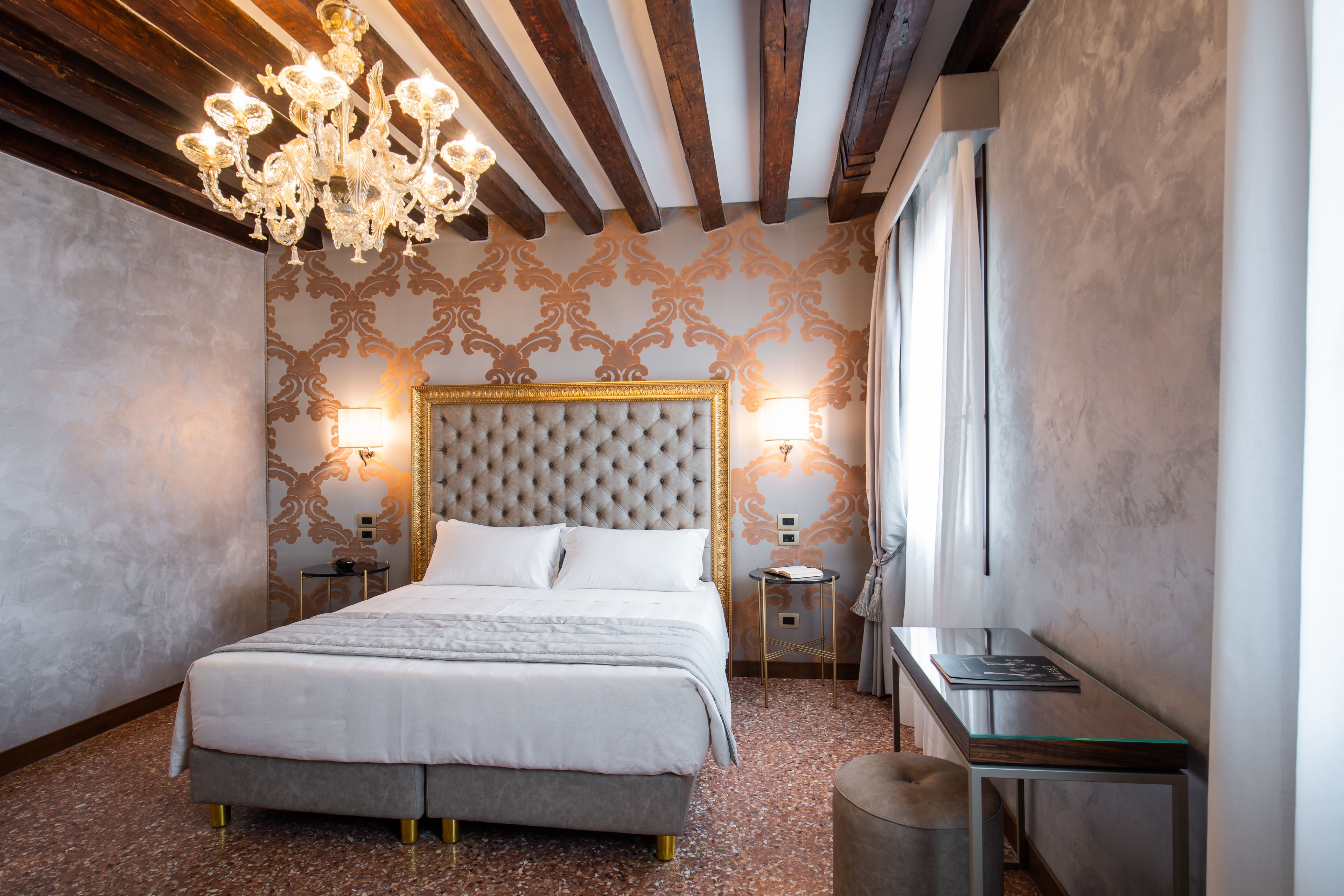 Quarto com cabeceira estofada, uma cama grande, um candelabro, uma mesa de cabeceira e um banquinho redondo.
