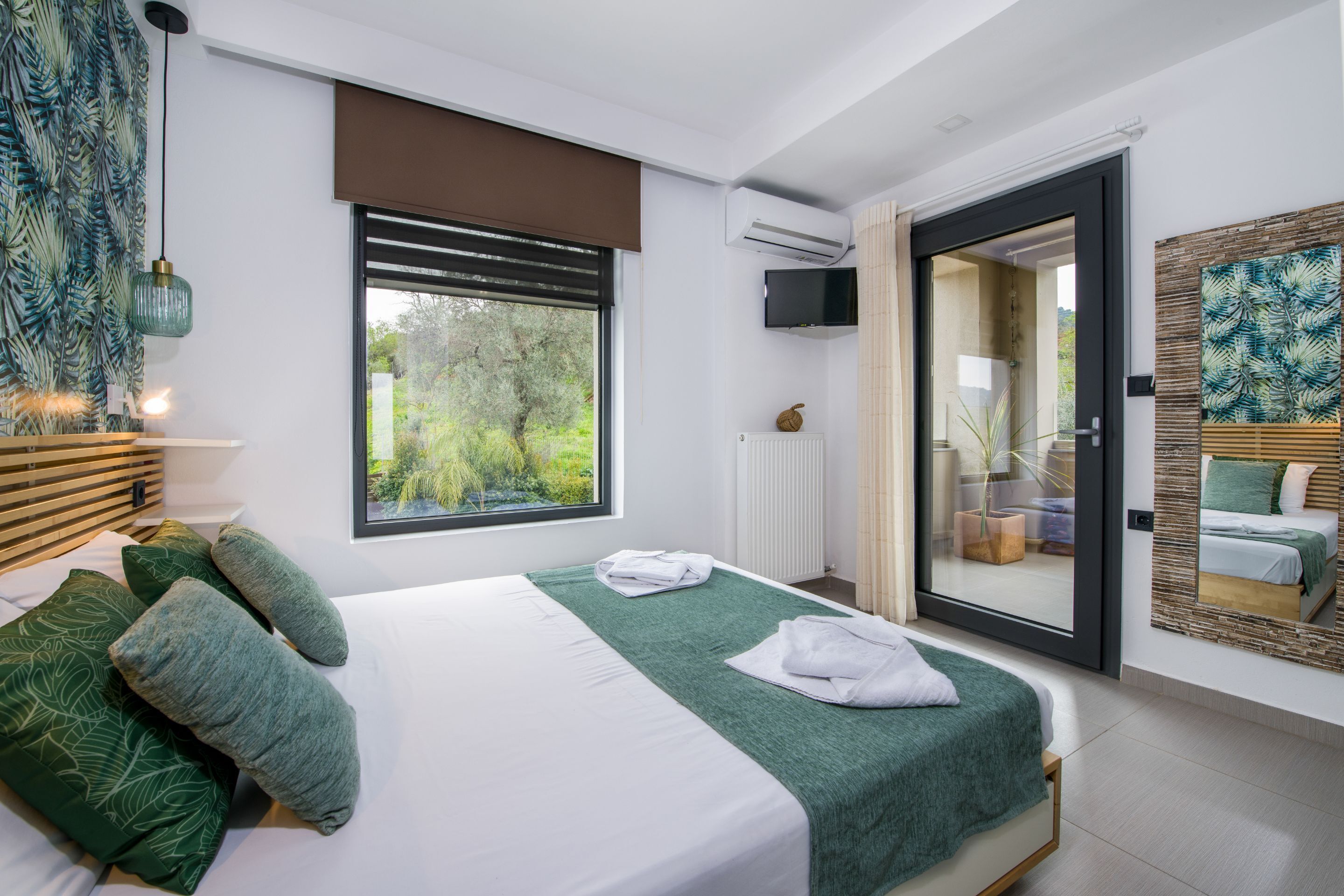 Quarto moderno com uma cama grande, almofadas verdes, vista para vegetação lá fora, porta de vidro deslizante e televisão montada na parede.