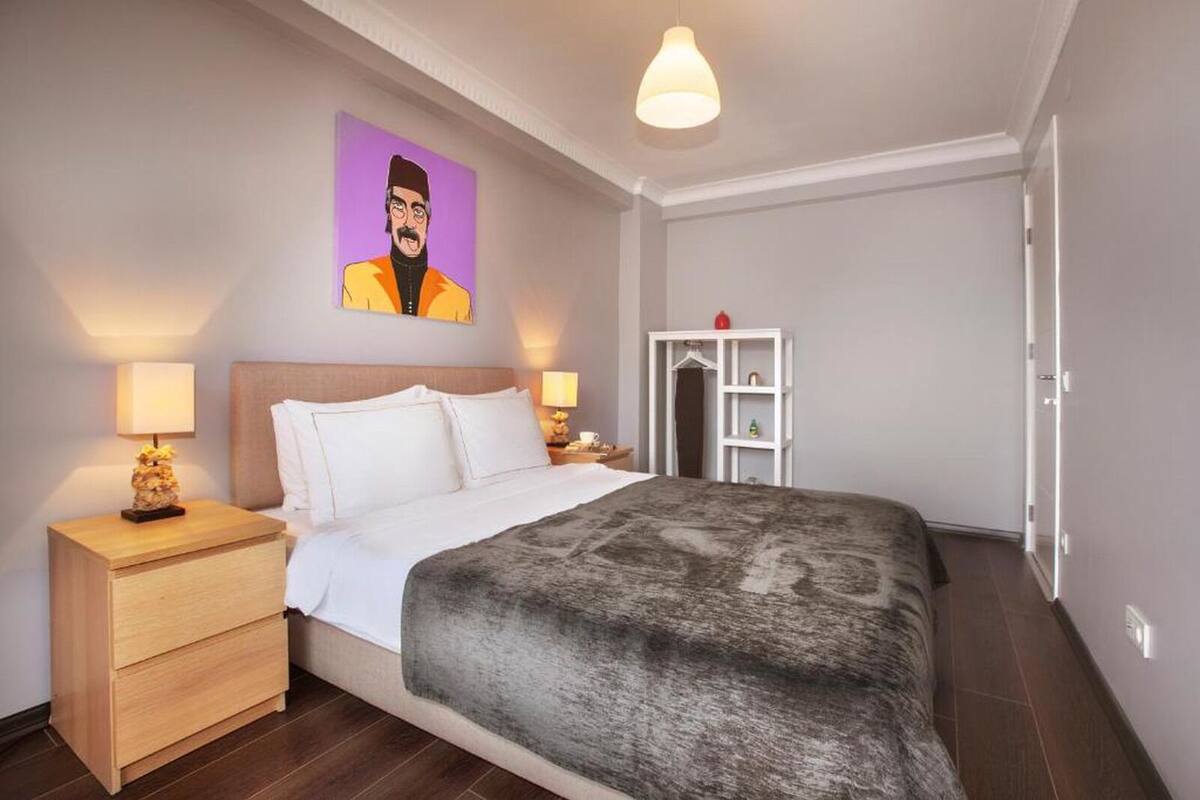 Quarto de hotel com uma cama, duas mesas de cabeceira com luminárias, um guarda-roupa e um quadro na parede.