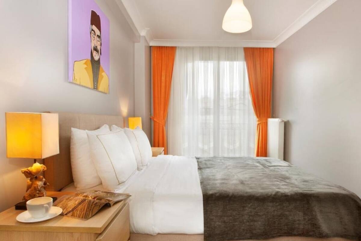 Quarto de hotel com uma cama, duas mesas de cabeceira com abajures, um quadro na parede e cortinas laranja.