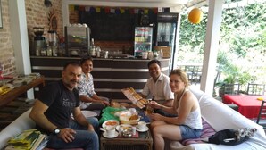 Um grupo de pessoas sentado à mesa, com comida e bebidas, uma máquina de café e uma geladeira ao fundo.