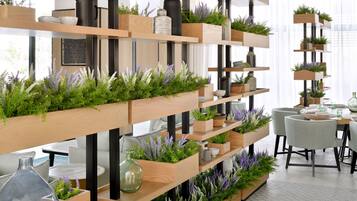 Interior moderno com prateleiras de madeira exibindo plantas e objetos decorativos, uma área de jantar com mesa e cadeiras redondas, e vista para o exterior por grandes janelas.