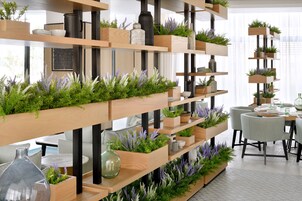 Interior moderno com prateleiras de madeira exibindo plantas e objetos decorativos, uma área de jantar com mesa e cadeiras redondas, e vista para o exterior por grandes janelas.