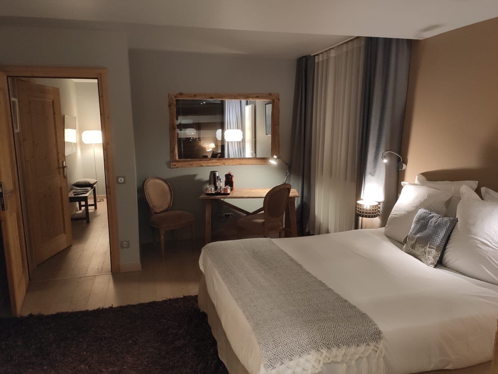 Quarto de hotel com uma cama grande, uma escrivaninha com cadeira, um espelho e uma janela com cortinas.