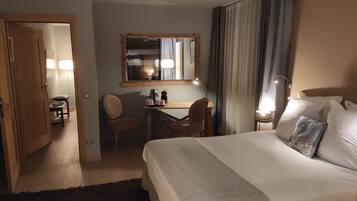 Quarto de hotel com uma cama grande, uma escrivaninha com cadeira, um espelho e uma janela com cortinas.