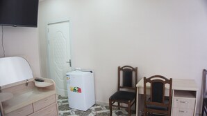 Um quarto pequeno e modesto, com um frigorífico branco, uma cadeira de madeira, uma escrivaninha de madeira e uma televisão na parede.