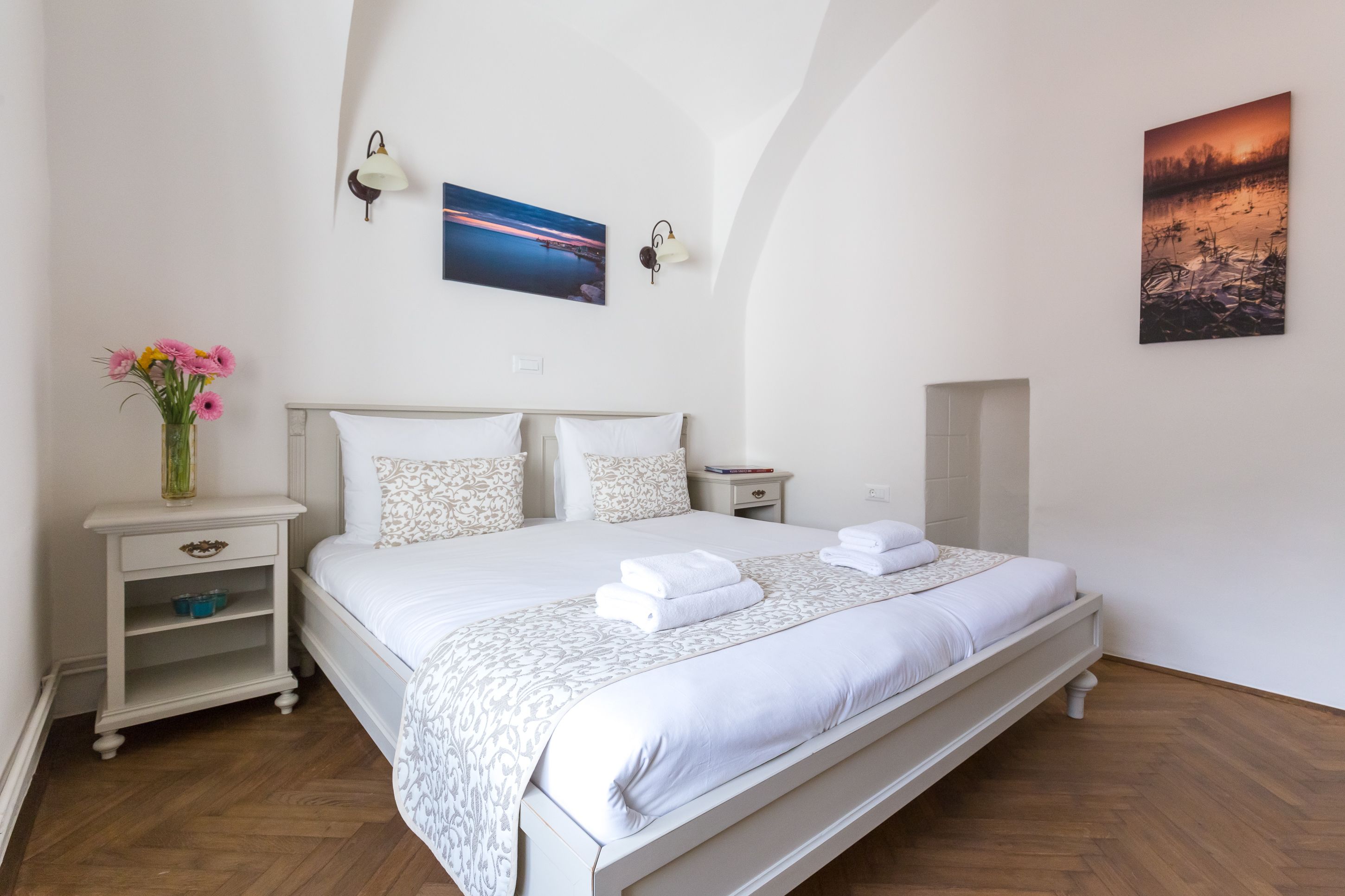 Quarto com cama grande, mesa de cabeceira, um vaso de flores e duas luminárias na parede.