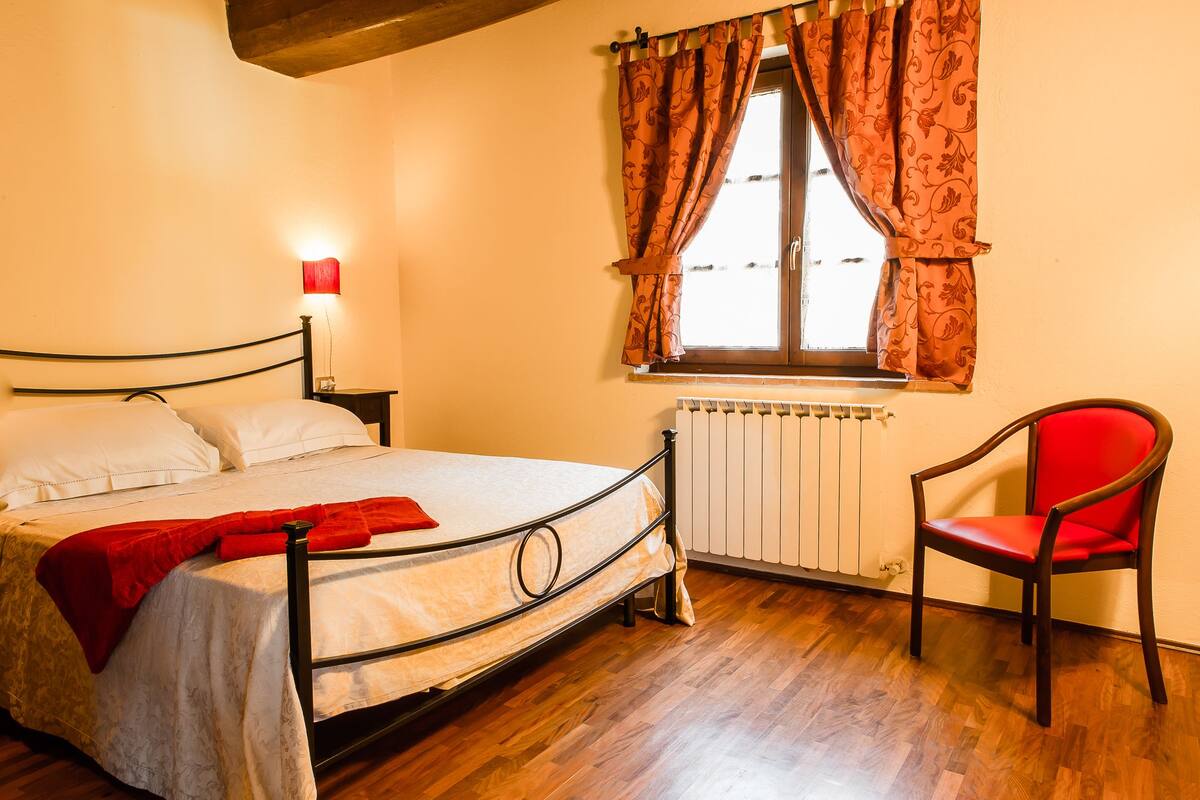 Quarto com cama, cadeira, janela com cortinas, radiador e luminária na parede.