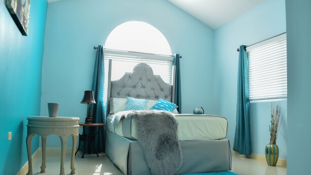 Quarto com cama, mesa de cabeceira, abajur, vaso e janela com cortinas.