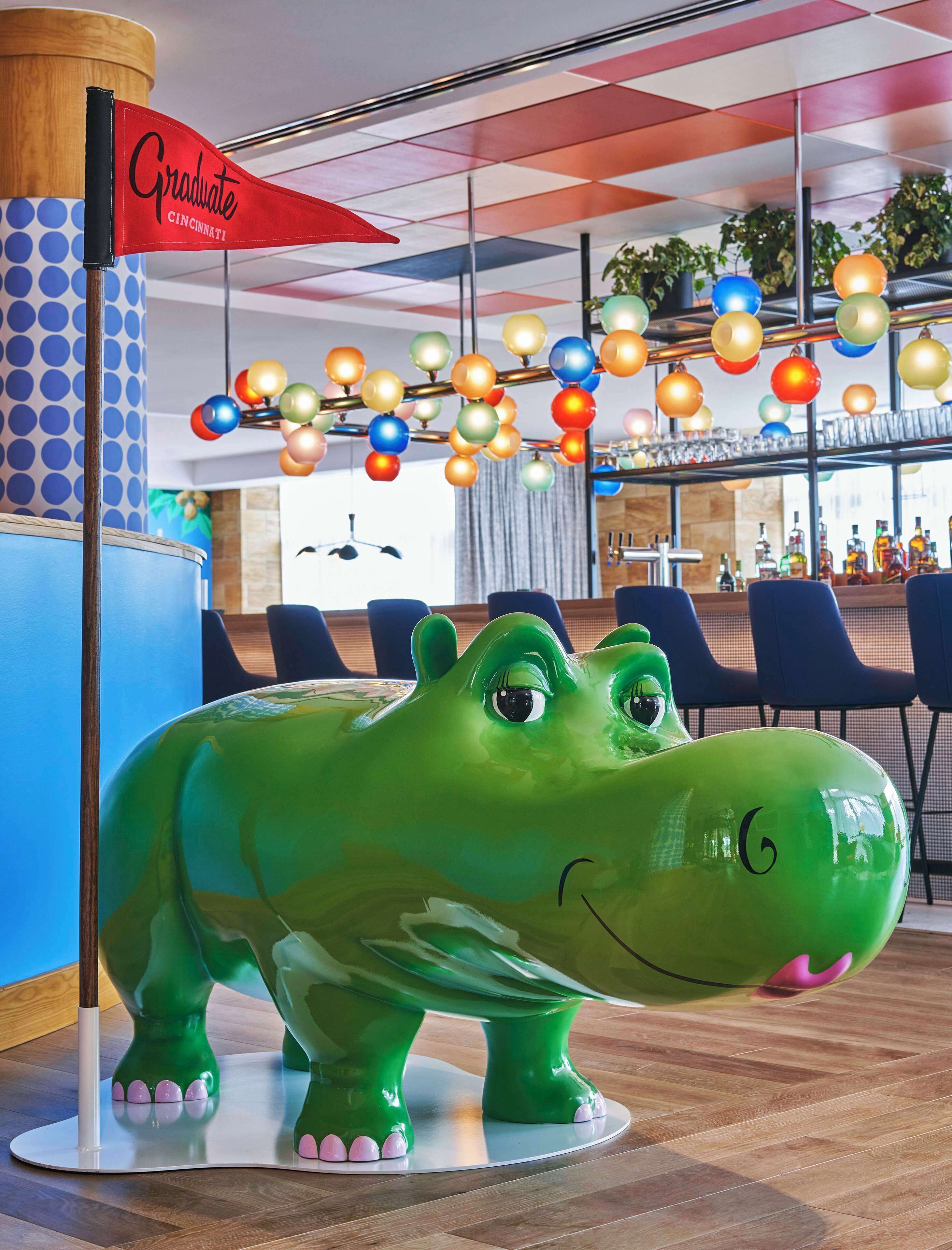 Uma estátua de hipopótamo verde de plástico em uma área de bar moderna, com uma bandeira vermelha 'Graduate Cincinnati', luzes pendentes coloridas e um balcão de bar com garrafas.
